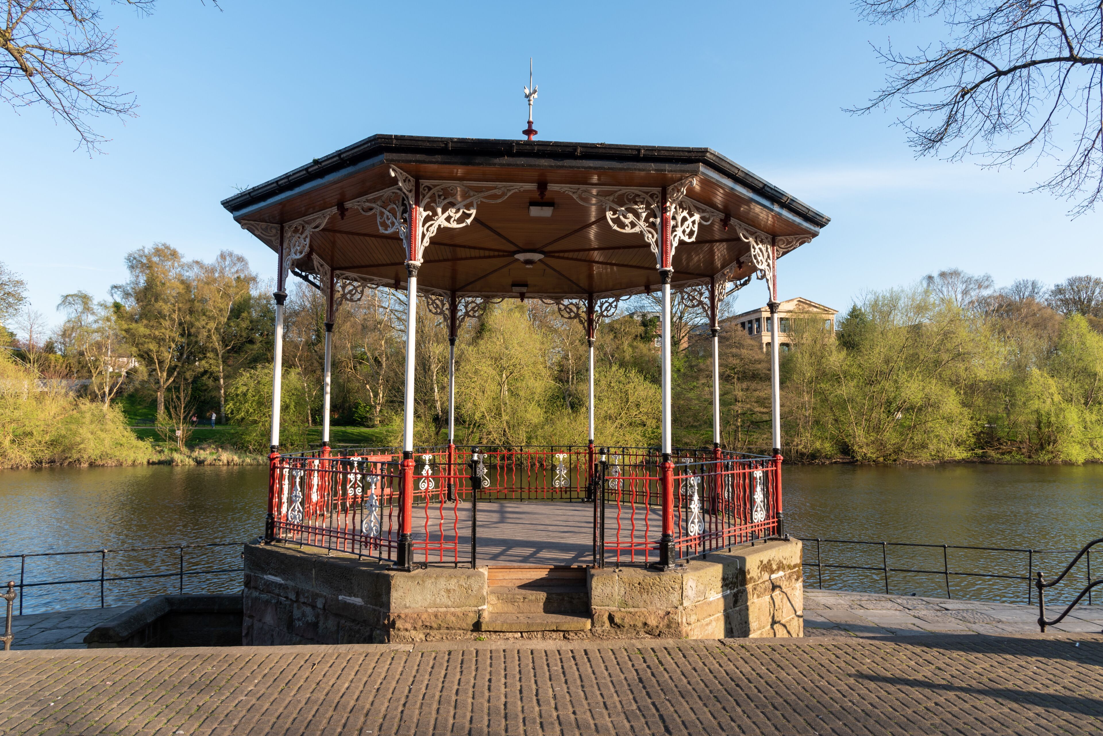 Um gazebo com detalhes metálicos ornamentados, localizado às margens de um rio calmo, cercado por árvores e um caminho de tijolos.