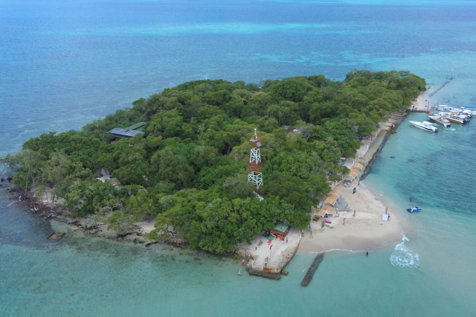 Vista aérea de uma ilha tropical com vegetação verde exuberante, uma praia de areia e várias embarcações atracadas perto da costa.