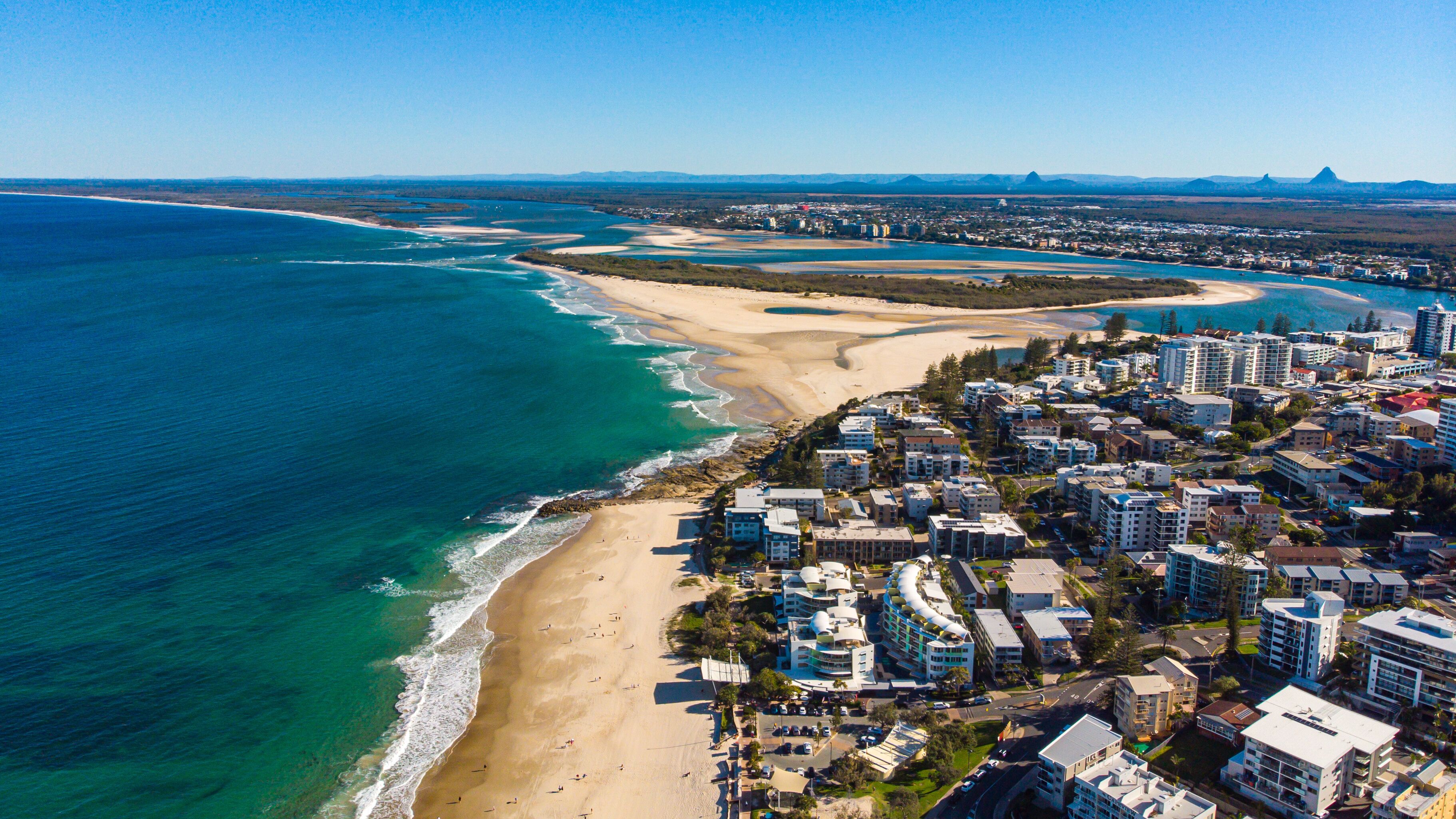 Paisagem costeira com praia, edifícios residenciais e uma cadeia de montanhas ao longe.
