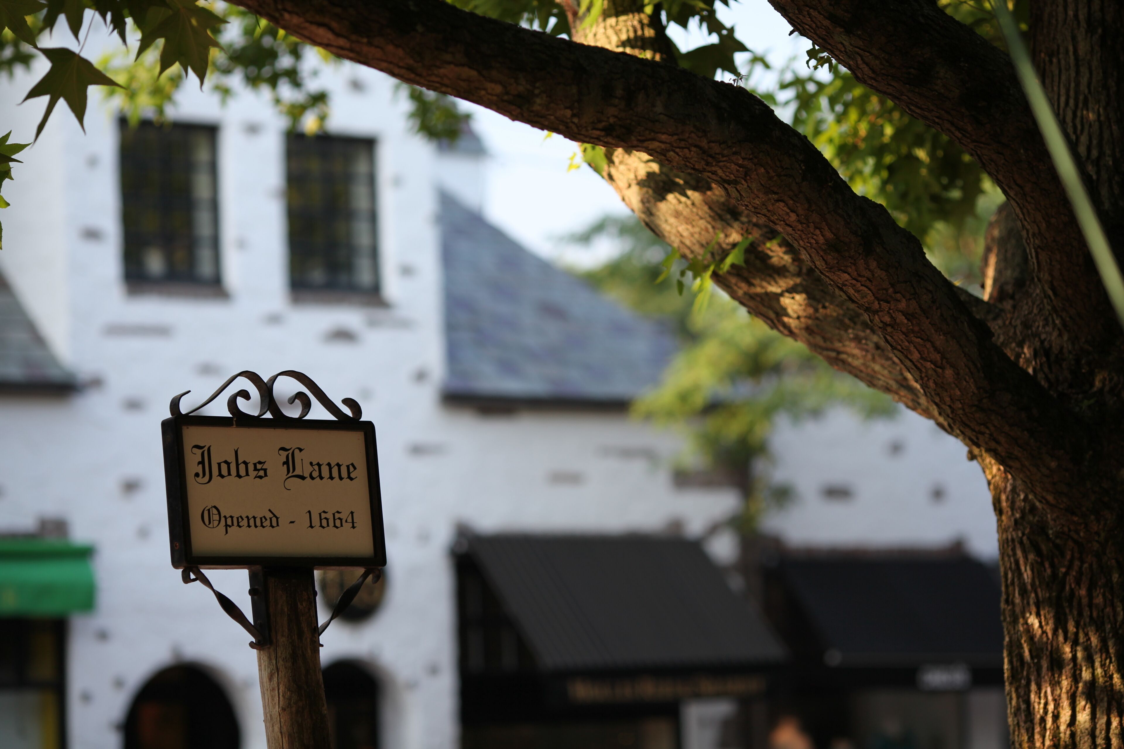 Uma placa de rua com a inscrição 'Jobs Lane Opened - 1664' em frente a um edifício com janelas em arco e telhado inclinado.