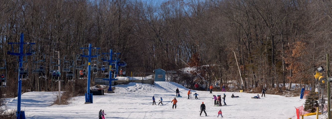 Pista de esqui coberta de neve com esquiadores e snowboarders, teleféricos e uma área arborizada ao fundo.