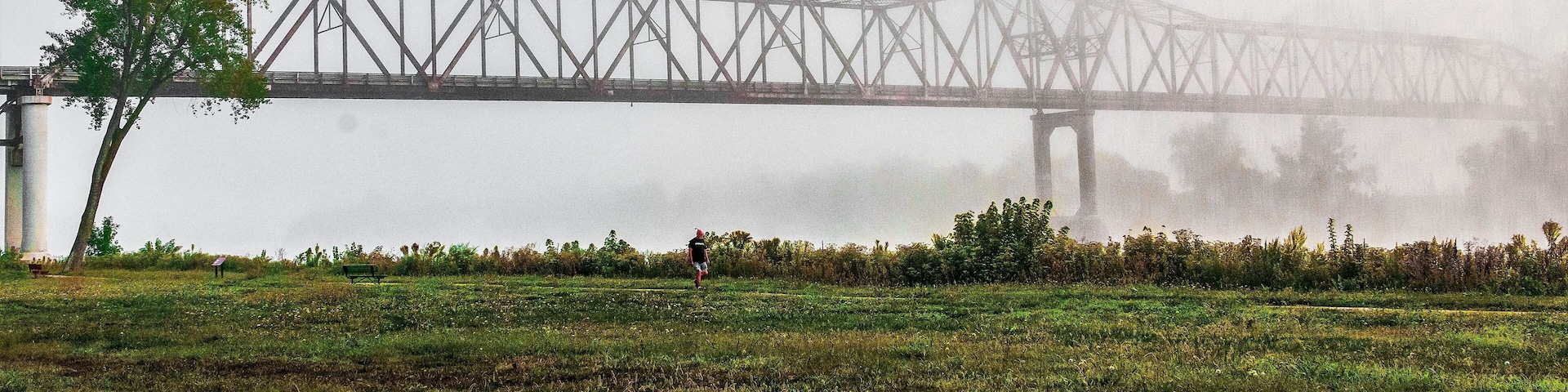 Uma ponte sobre a água, cercada por um campo gramado com uma única árvore e uma pessoa caminhando.