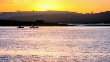 Um pôr do sol sobre águas calmas, com barcos e uma colina ao longe.