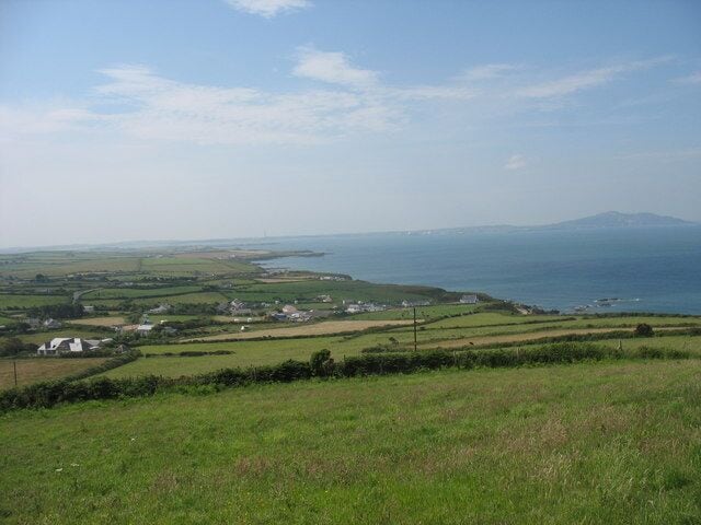 Paisagem costeira com colinas onduladas, casas dispersas e vista distante do mar e de uma montanha.