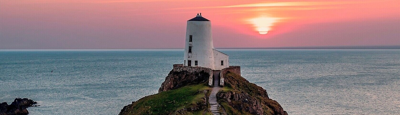 Um farol numa formação rochosa com um caminho que o leva a um mirante, com vista para o mar ao pôr do sol.