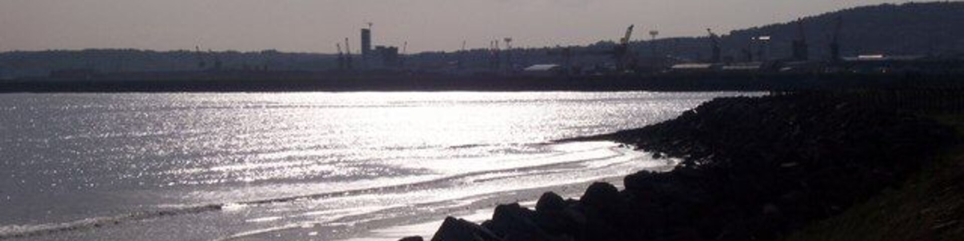 Paisagem costeira com linha de costa rochosa, águas calmas refletindo o sol e estruturas industriais ao longe.