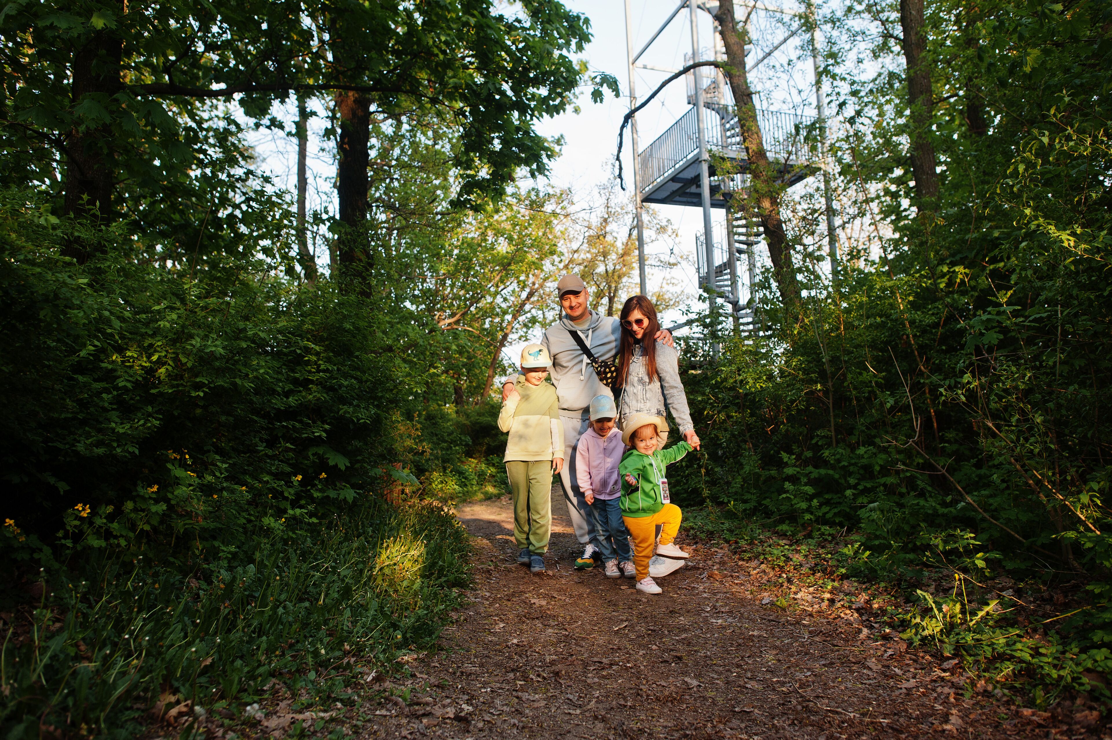 Uma família caminhando por uma trilha na floresta, com vegetação densa e uma estrutura de metal ao fundo.