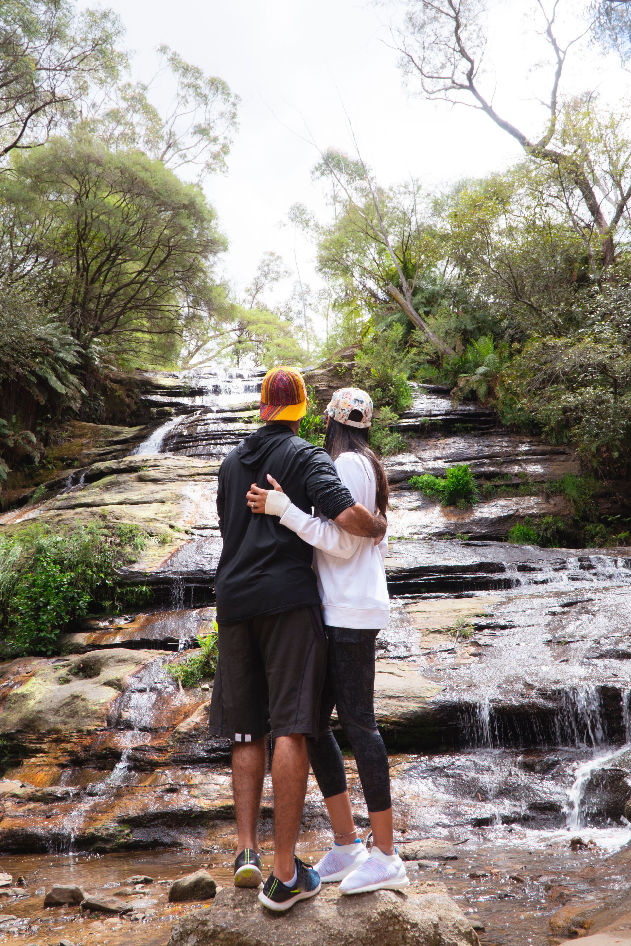 Um casal em pé sobre uma cachoeira rochosa, com vegetação exuberante ao redor.