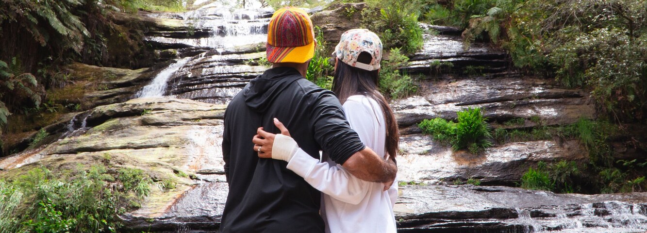 Um casal em pé sobre uma cachoeira rochosa, com vegetação exuberante ao redor.