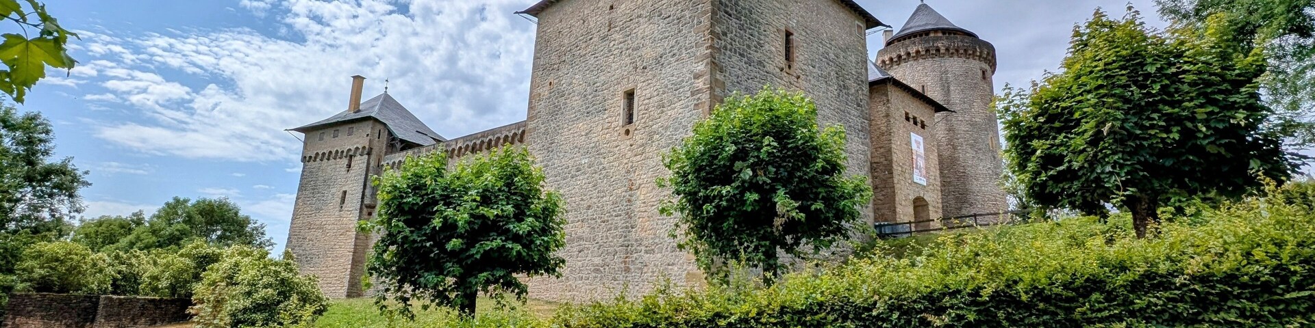 Um castelo de pedra histórico com duas torres, rodeado por vegetação exuberante e um céu limpo.