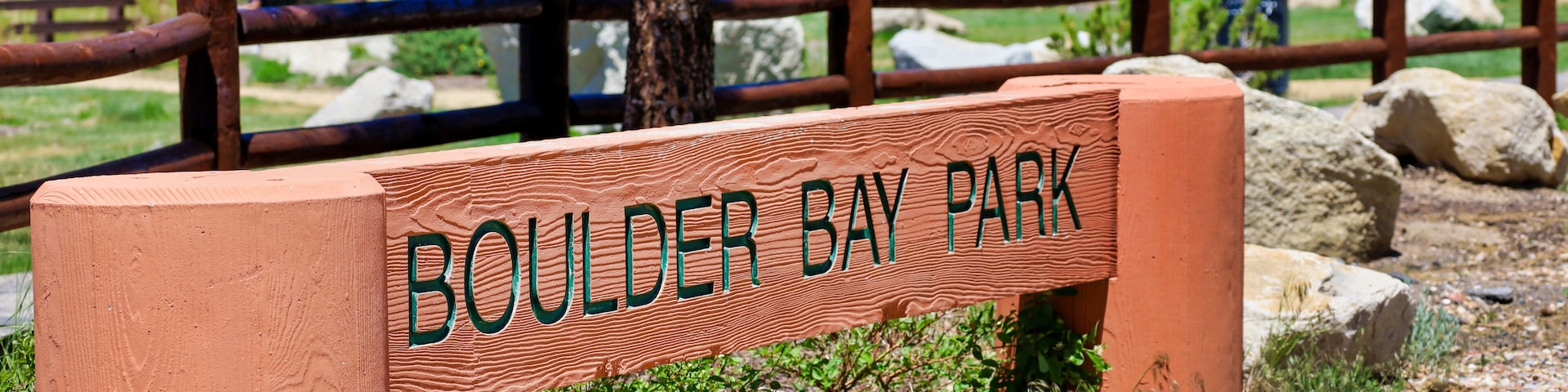 Uma placa de madeira com a inscrição 'BOULDER BAY PARK', rodeada por pedras e vegetação.