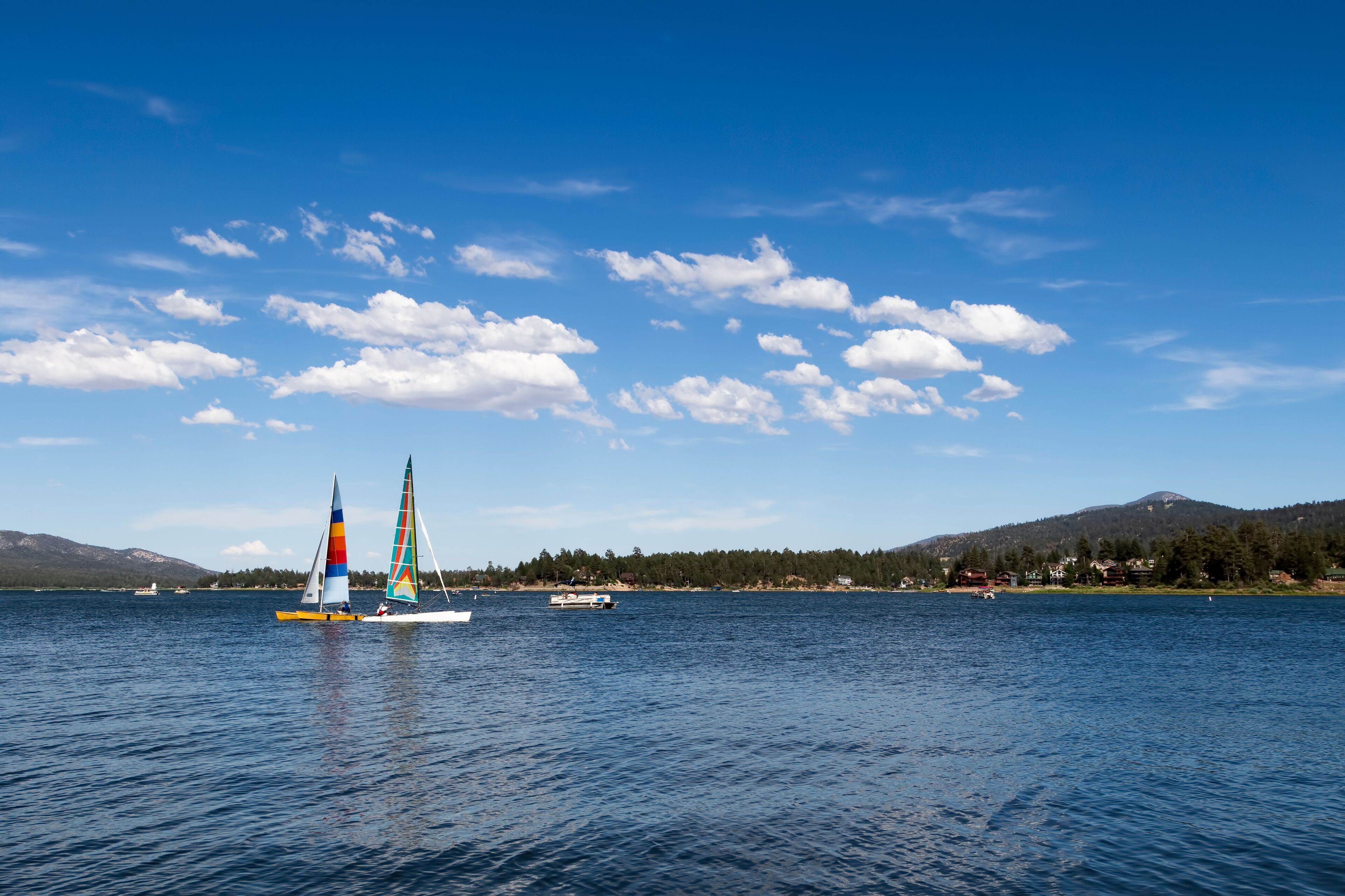 Um lago tranquilo com barcos à vela, uma linha de costa distante com casas e uma cadeia de montanhas sob um céu limpo.