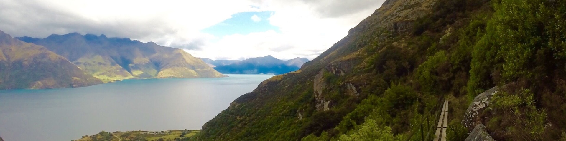 Uma ponte suspensa sobre um vale, com um lago e montanhas ao fundo.