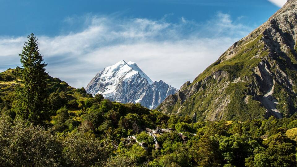Paisagem montanhosa com um pico nevado imponente, floresta verde densa e terreno acidentado.