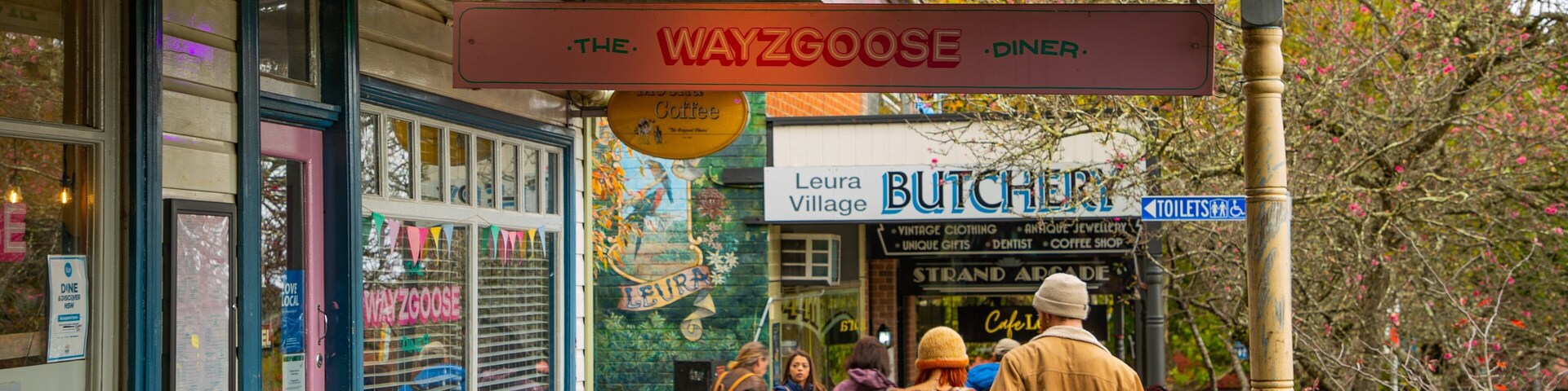 Cena de rua com uma cafeteria chamada 'The Wayzgoose Diner' e uma açougue chamada 'Leura Village Butcher'.