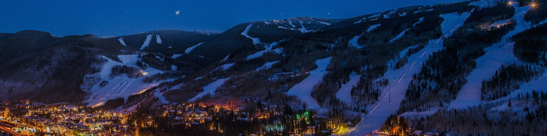 Uma cidade nas montanhas coberta de neve à noite, com edifícios iluminados e uma lua crescente no céu.