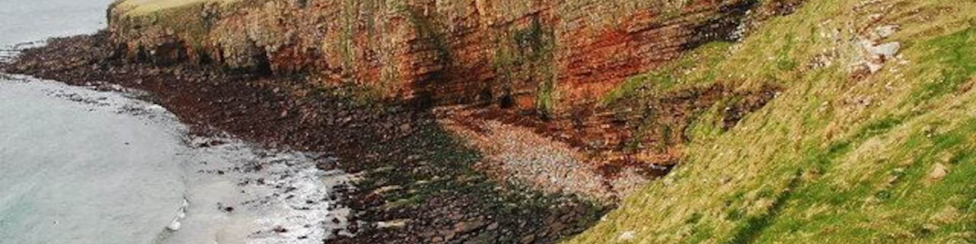 Um penhasco costeiro com camadas de rocha vermelhas e verdes, uma encosta gramada e uma praia rochosa.