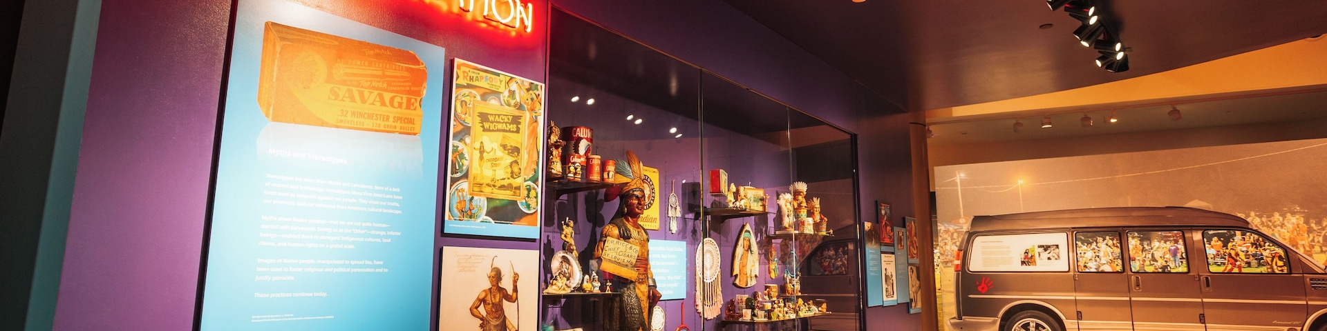 Uma exposição de museu com um letreiro de neon que diz 'MIS-REPRESENTAÇÃO', vitrines com livros e artefatos, e uma van com imagens nas laterais.