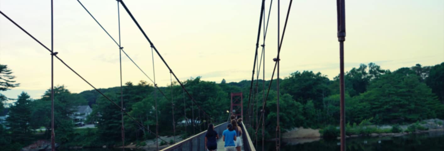 Uma ponte suspensa sobre águas calmas, com uma paisagem arborizada ao fundo.