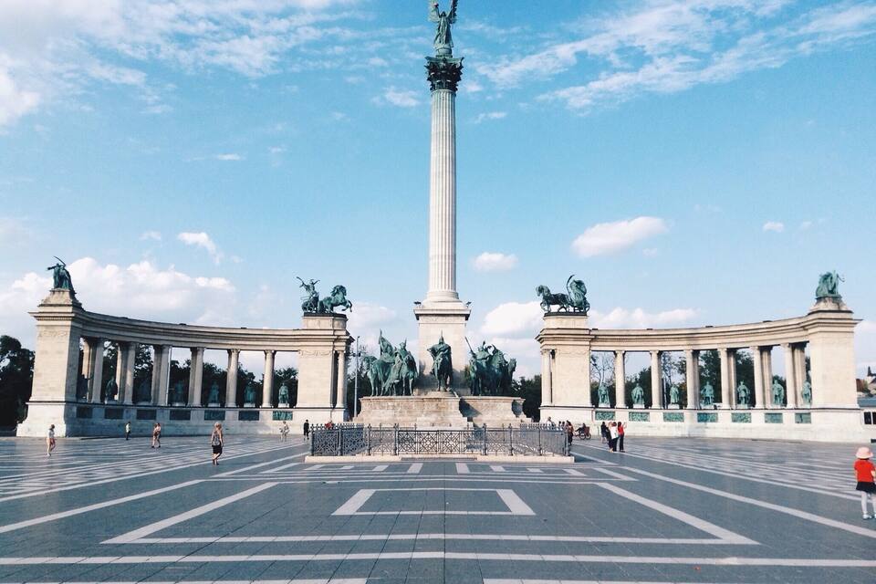Uma grande praça aberta com um monumento central, ladeada por dois edifícios com colunas e estátuas.