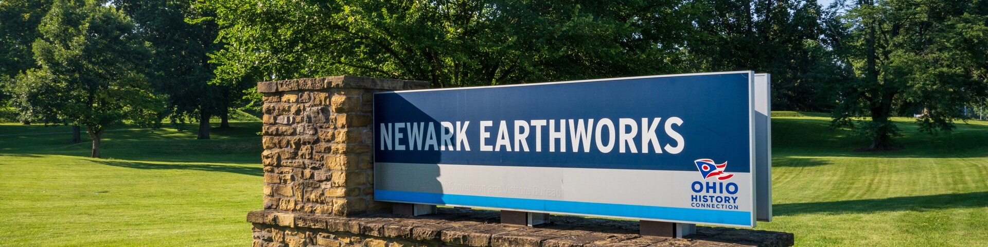 Um sinal de pedra com o texto 'Newark Earthworks' e 'Ohio History Connection', rodeado por um campo gramado e árvores.