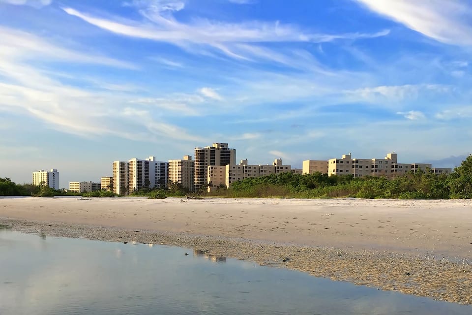Uma praia com mar calmo, céu limpo e edifícios ao fundo.