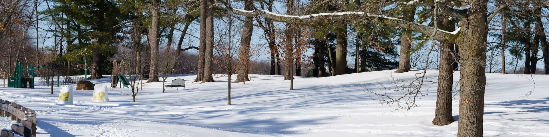 Um parque coberto de neve com bancos de madeira, árvores e uma cerca de madeira.