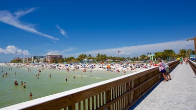 Praia movimentada com pessoas nadando e tomando sol, um calçadão de madeira e palmeiras.