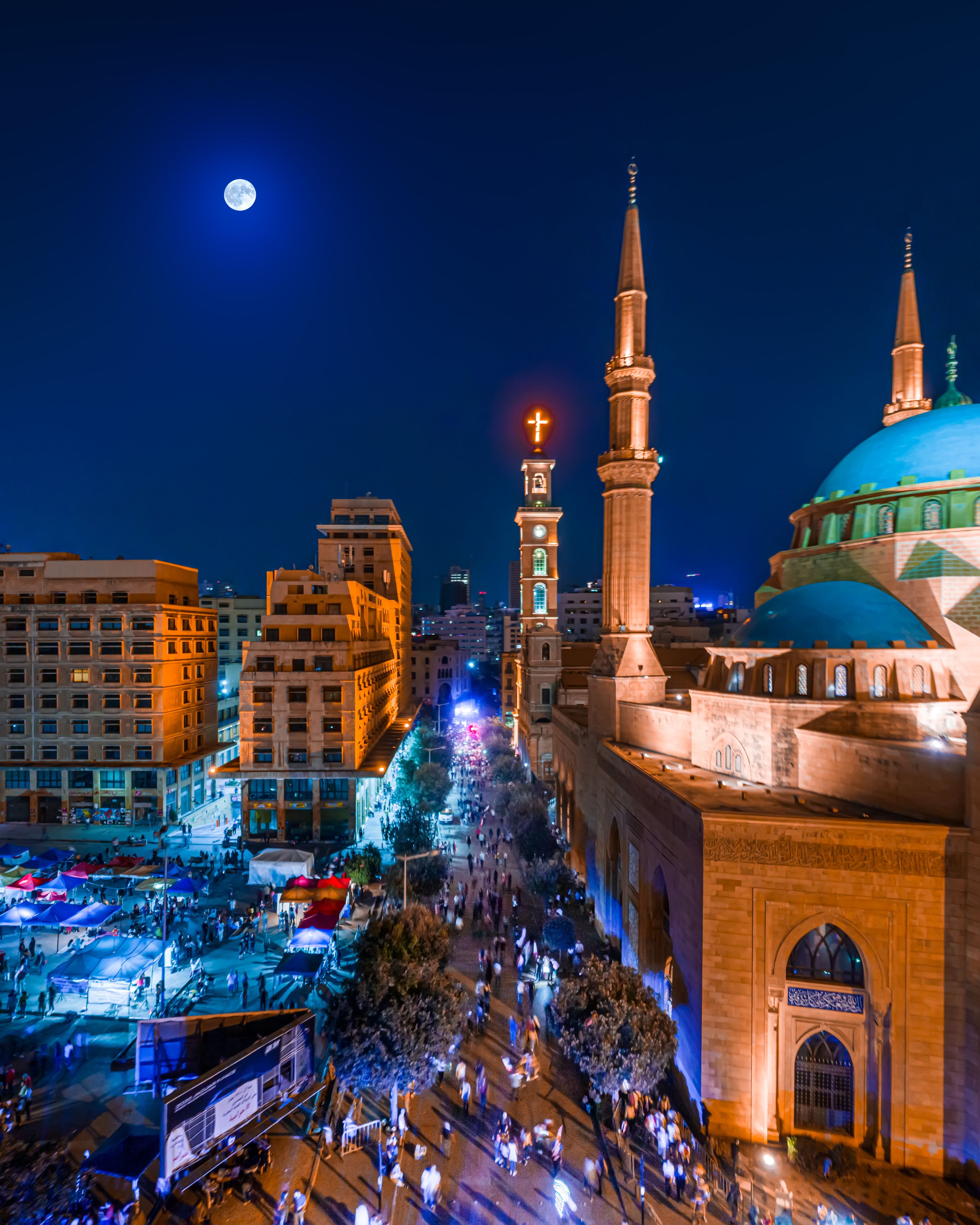 Visão urbana noturna com uma mesquita em destaque, com duas torres e uma cúpula grande, edifícios iluminados e uma lua visível no céu.