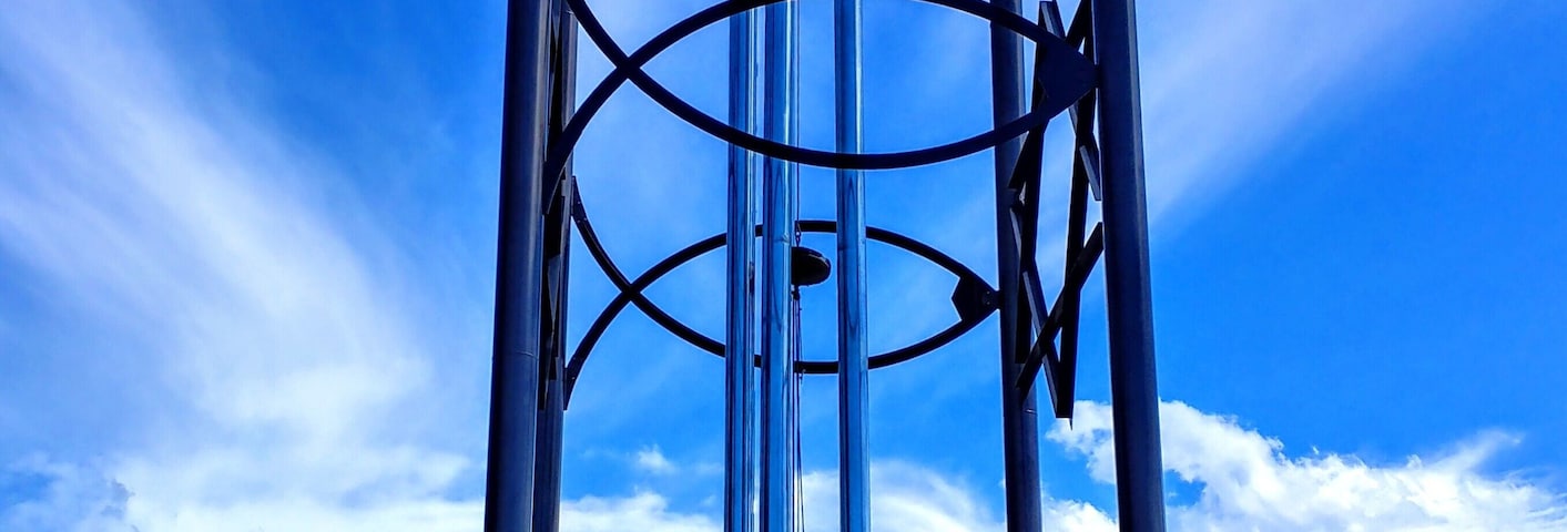 Escultura metálica alta, com base circular e braços entrelaçados, destacando-se contra um céu azul límpido com nuvens.