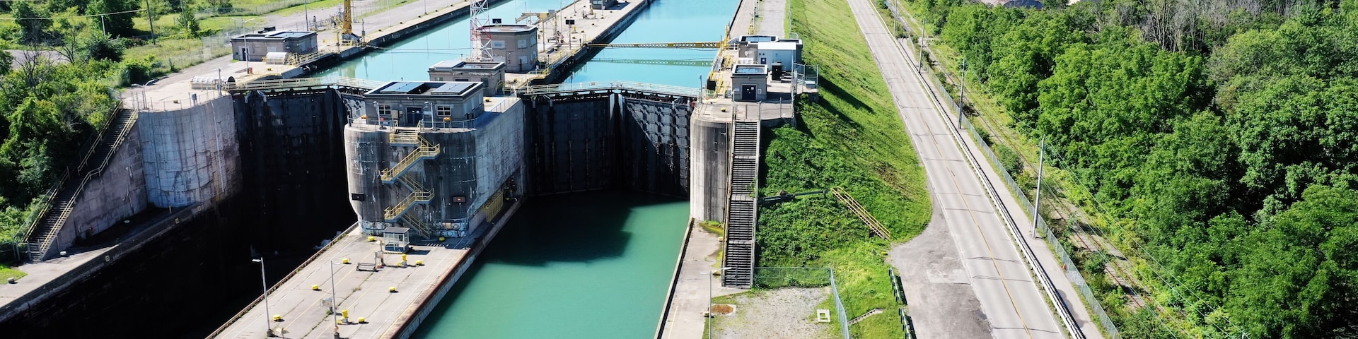 Uma barragem com canais de água, uma estrada ao lado e uma área arborizada ao fundo.