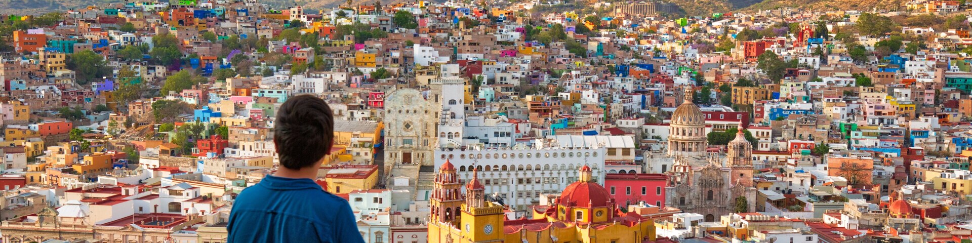 Uma pessoa sentada em um parapeito, com vista para uma paisagem urbana densamente povoada, com edifícios coloridos e uma igreja com cúpula vermelha em destaque.