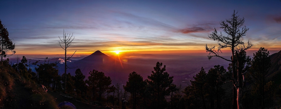 Montanha ao pôr do sol, com céu claro e silhuetas de árvores.