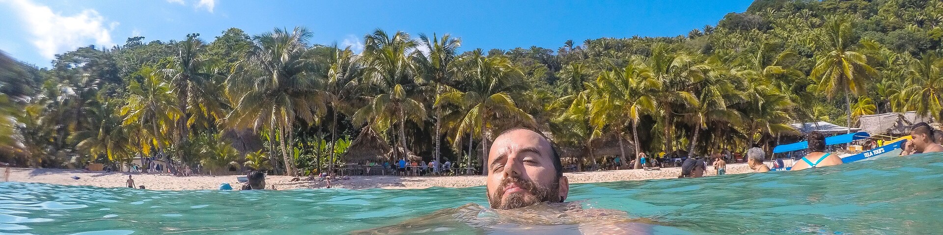 Uma pessoa nada em águas turquesa cristalinas, com uma praia e palmeiras ao fundo.
