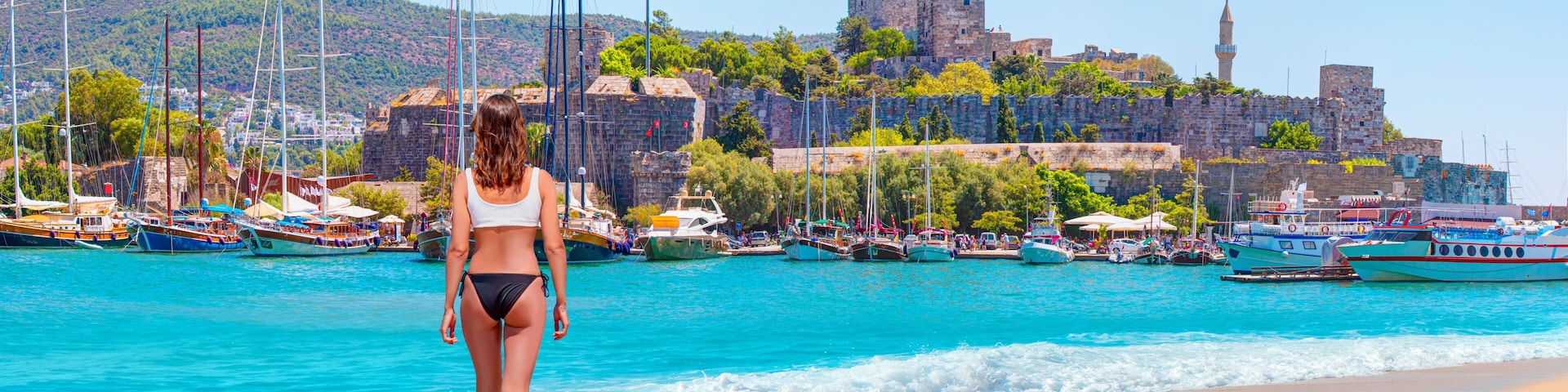 Uma praia com uma mulher de biquíni caminhando em direção à água, um castelo ao fundo e vários barcos atracados na costa.