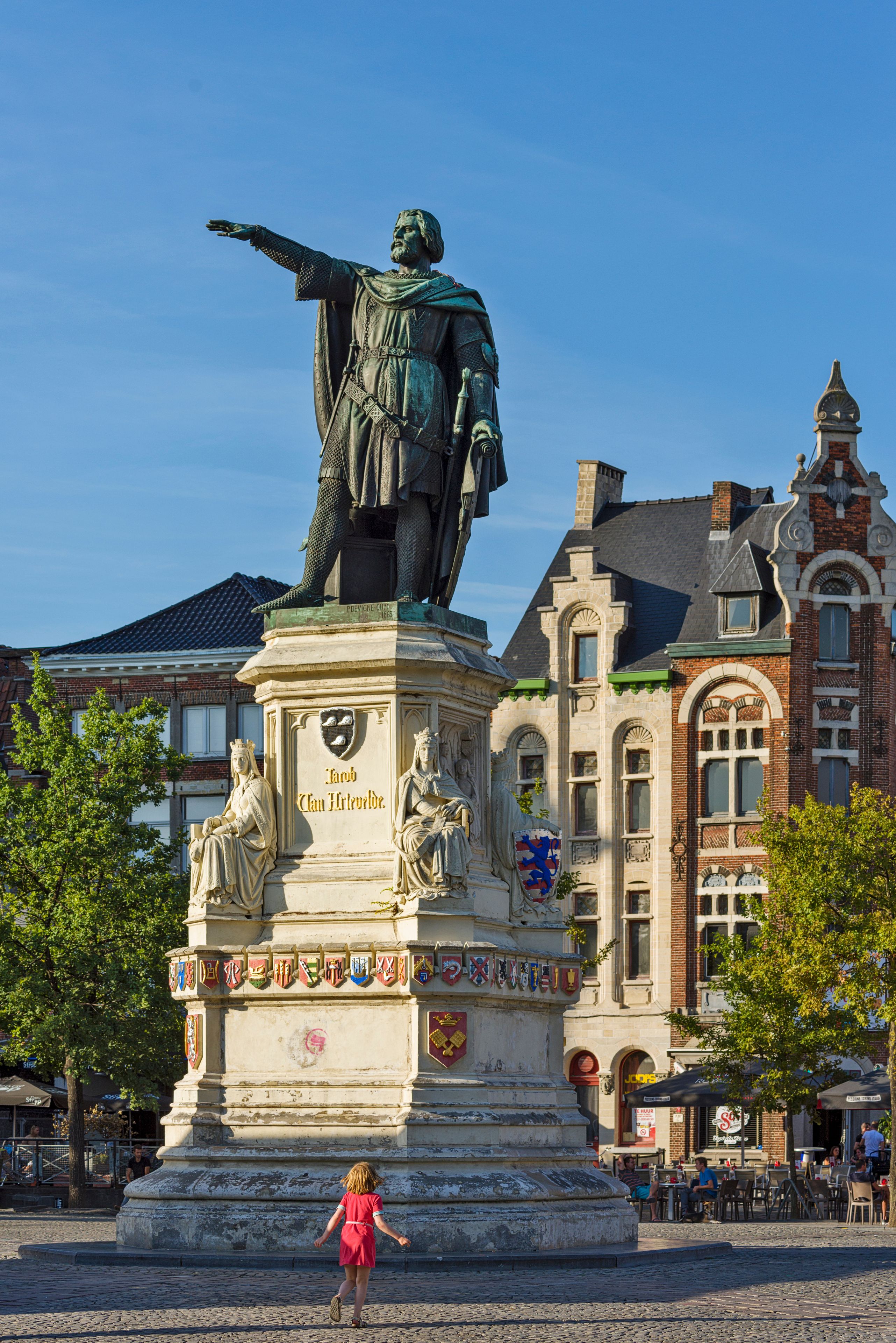 Estátua de uma figura histórica com o braço levantado, em um pedestal com brasões, em frente a um edifício com torre do relógio.