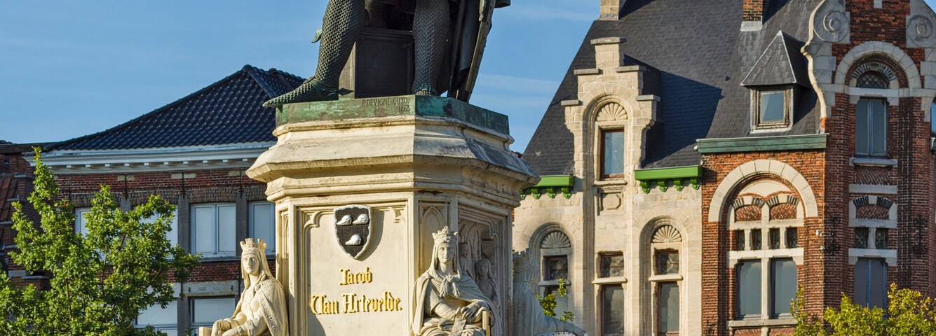 Estátua de uma figura histórica com o braço levantado, em um pedestal com brasões, em frente a um edifício com torre do relógio.