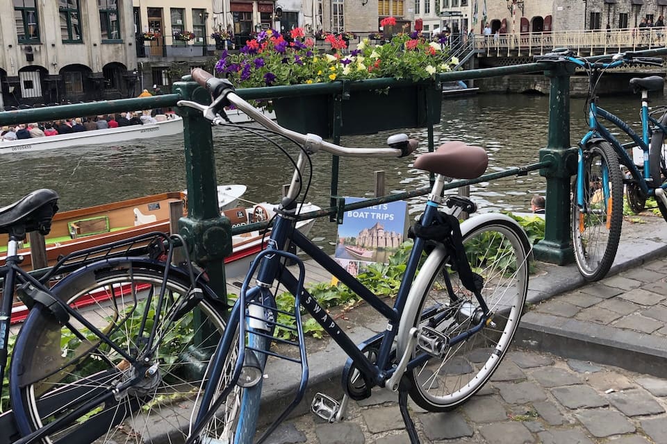 Uma bicicleta estacionada junto a um canal, com edifícios históricos e caixas de flores ao fundo.