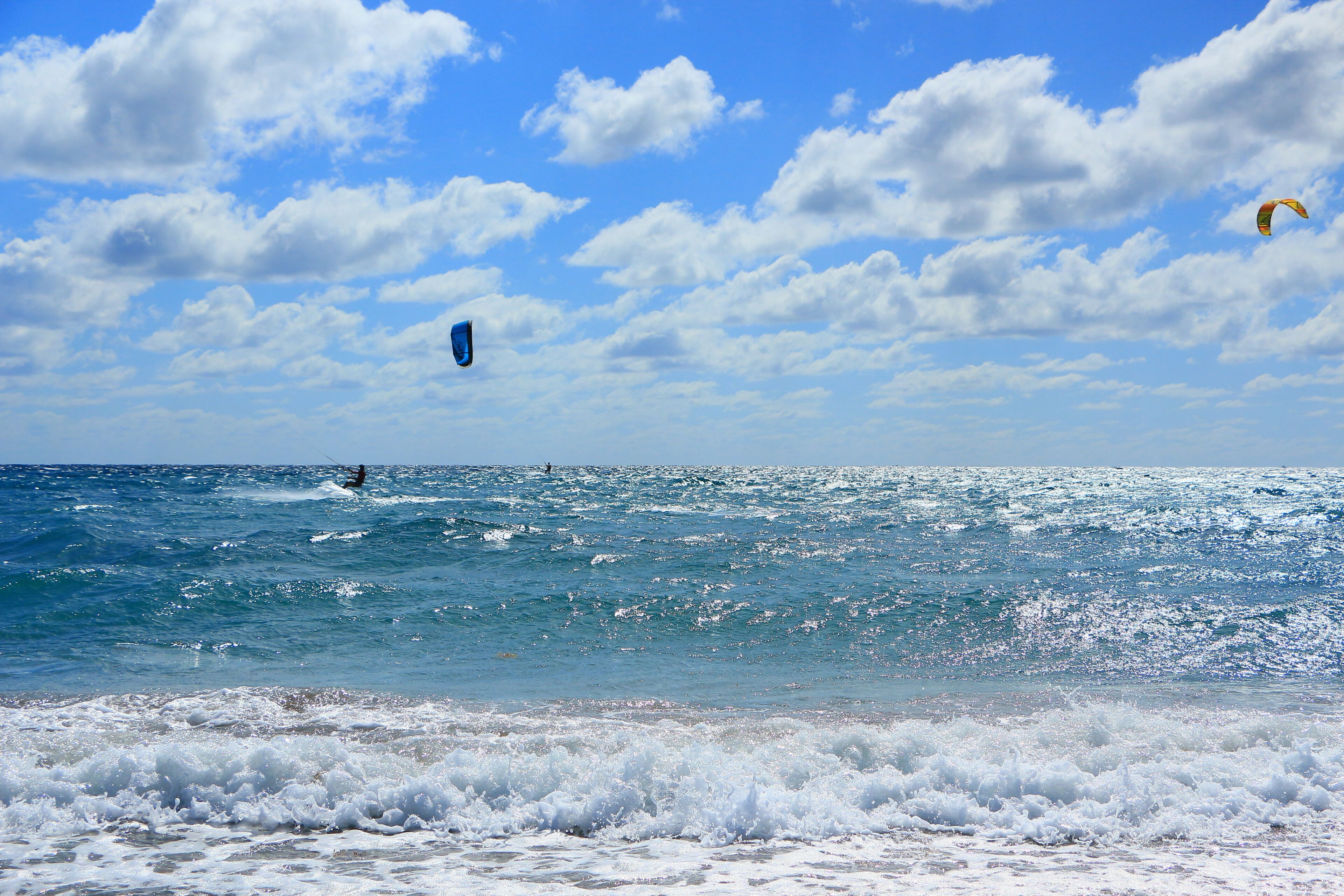 Praia com ondas, céu claro e uma pessoa praticando kitesurf.