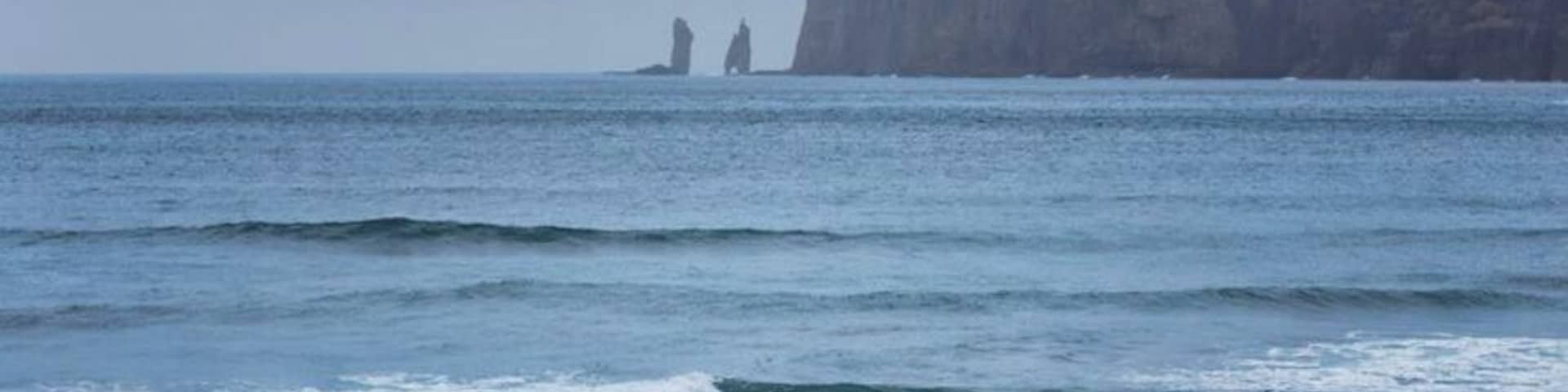 Uma praia com duas pessoas na água, ondas se aproximando da costa e um grande penhasco ao fundo.