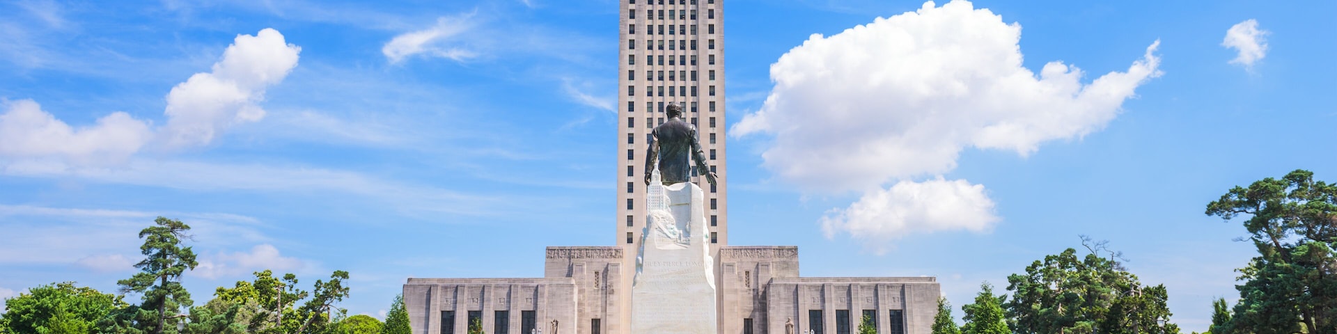 Um edifício alto com uma estátua na frente, rodeado por jardins bem cuidados e árvores.