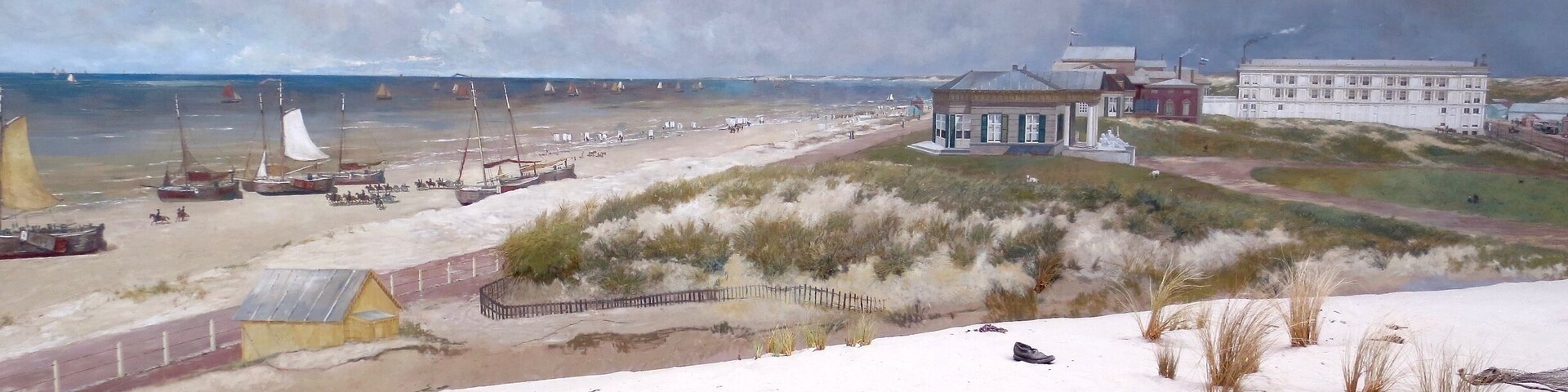 Uma praia com barcos, uma área arenosa com dunas e edifícios ao fundo.