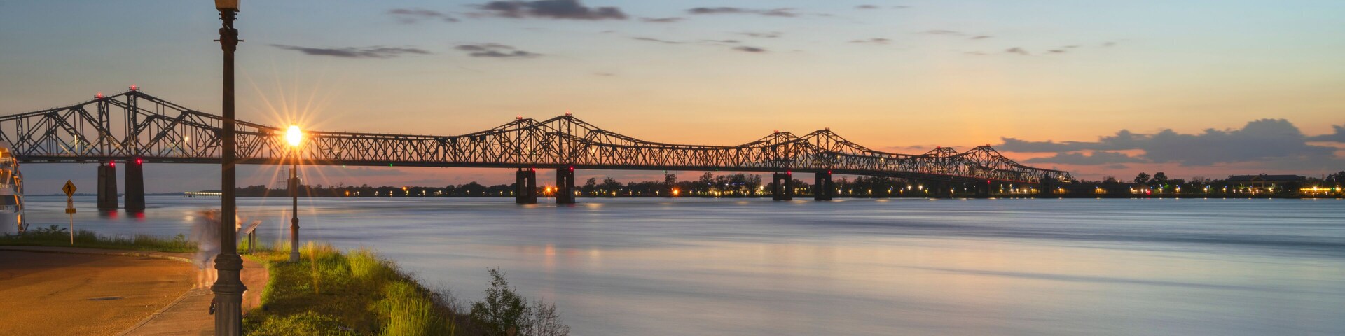 Pôr do sol sobre um rio com uma ponte, um poste de luz e uma margem gramada.