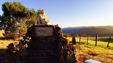 Estátua de mulher segurando uma criança em pedestal de pedra, com uma placa, em paisagem rural com árvores e colinas.