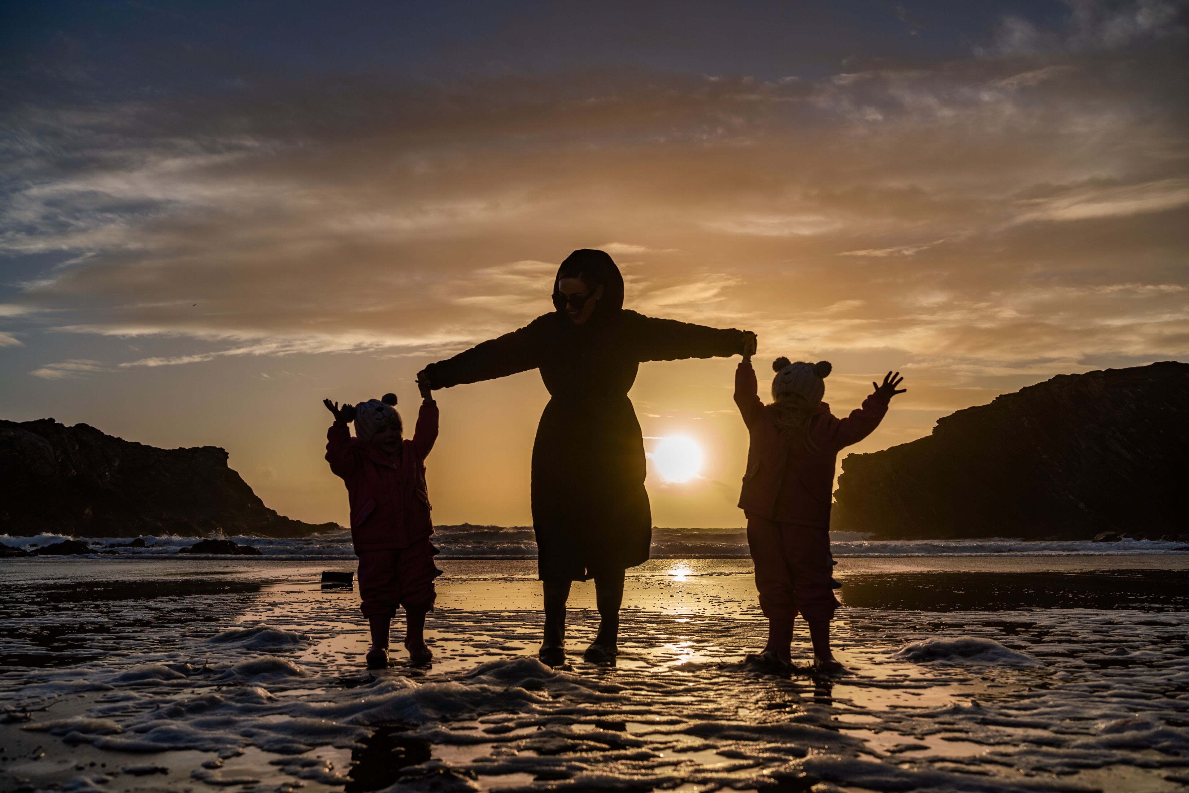 Silhueta de uma pessoa e duas crianças contra um pôr do sol numa praia com falésias rochosas ao fundo.