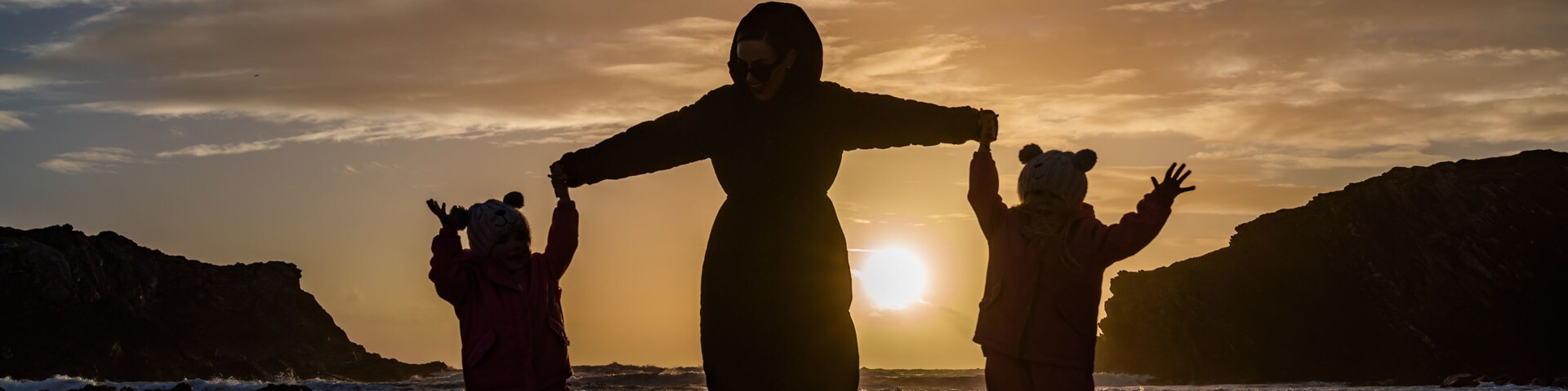 Silhueta de uma pessoa e duas crianças contra um pôr do sol numa praia com falésias rochosas ao fundo.