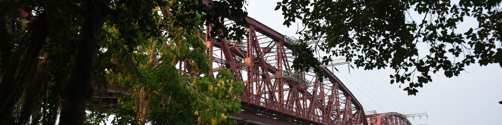 Uma grande ponte de aço vermelha atravessa um rio, cercada por vegetação exuberante e árvores.