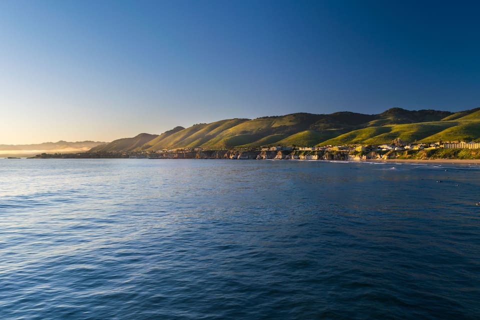 Paisagem costeira com céu claro, mar calmo e colinas ondulantes com edifícios ao longo da costa.