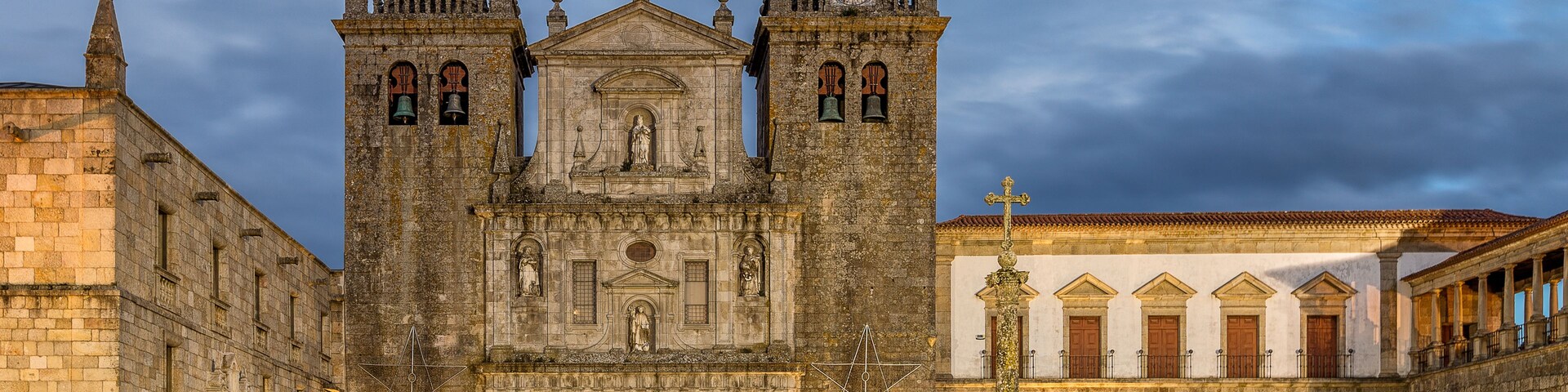 Uma igreja histórica com uma torre central e duas torres menores, rodeada por um pátio com uma fonte.