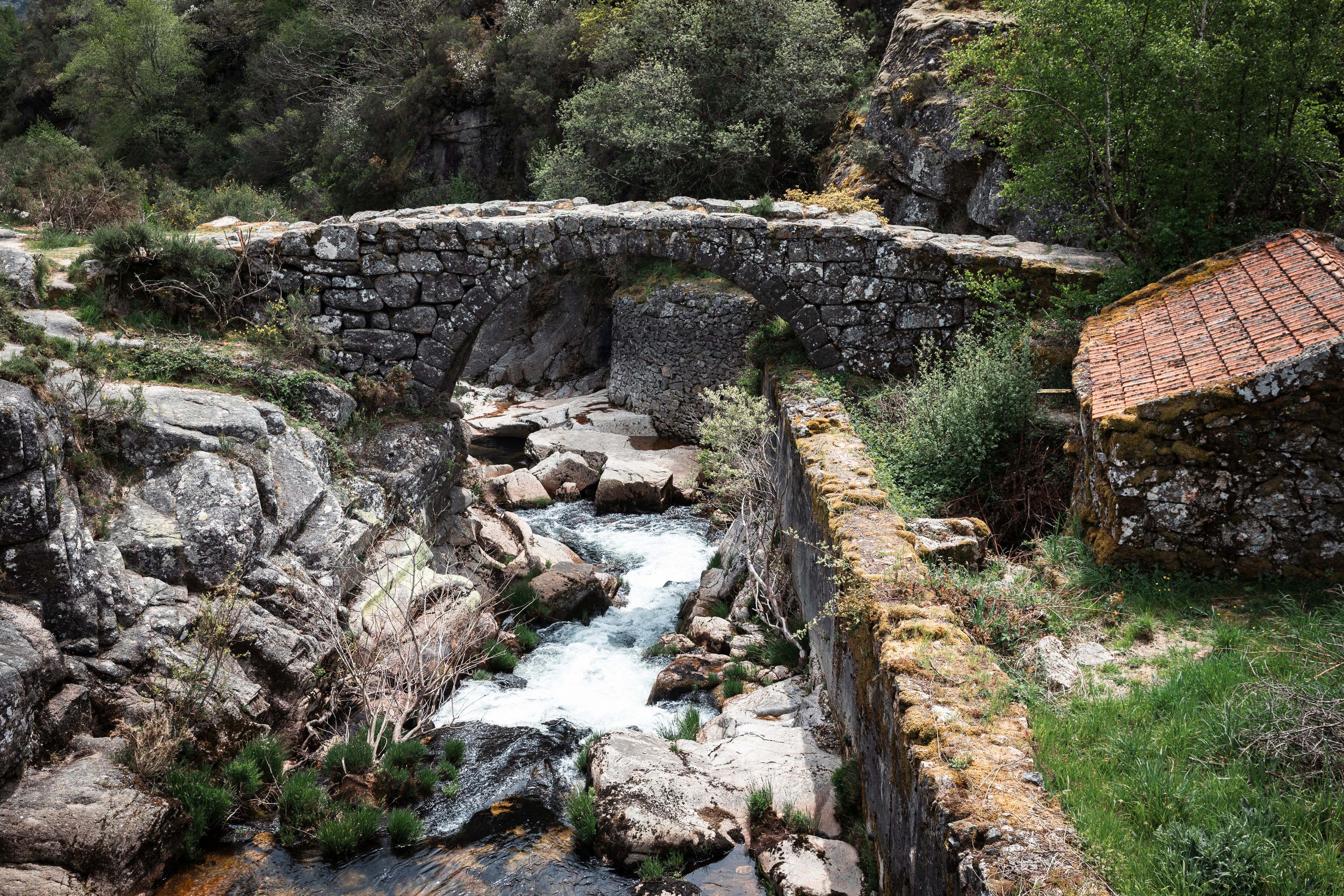 Uma ponte de pedra sobre um rio corrente, rodeada por terreno rochoso e vegetação.